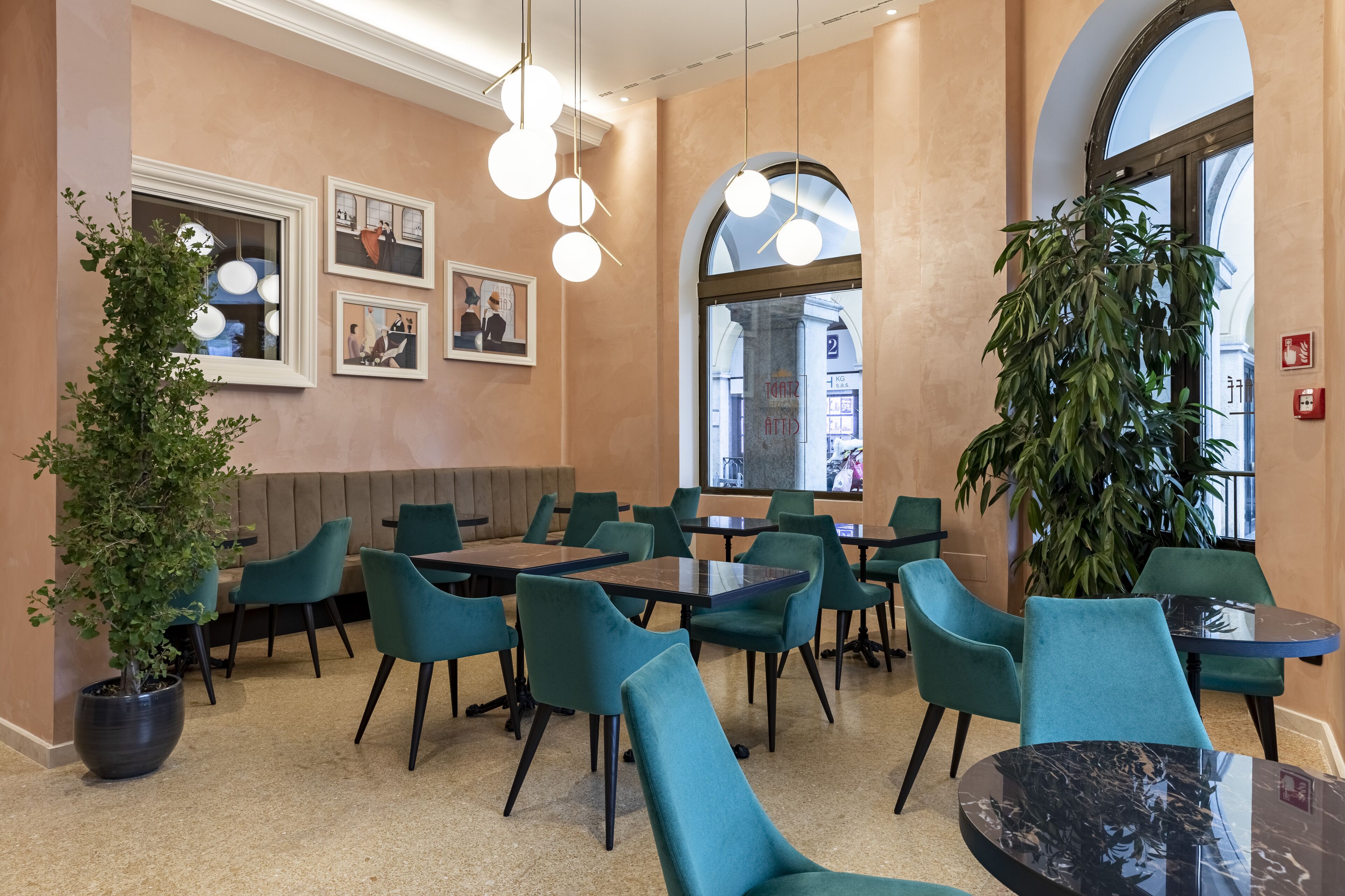 Um restaurante com cadeiras cor de verde-água e mesas pretas, uma grande planta em vaso e quadros nas paredes.