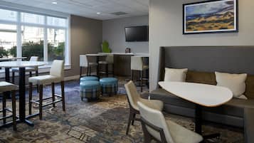 Um lobby de hotel com uma mesa redonda, banquetas de bar, um sofá, pufes, uma TV de tela plana e um quadro na parede.