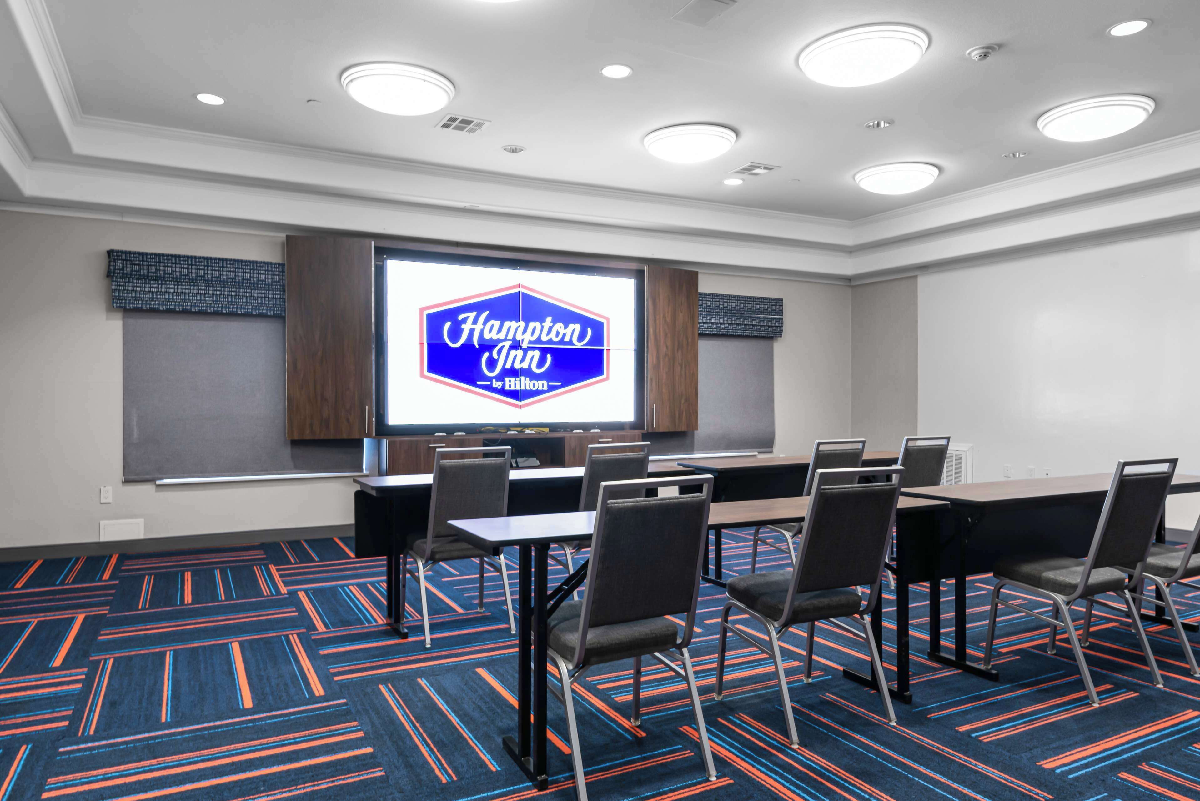 Sala de conferência com uma tela grande exibindo o logotipo do Hampton Inn, uma mesa comprida com cadeiras e um tapete estampado.