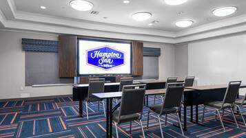 Sala de conferência com uma tela grande exibindo o logotipo do Hampton Inn, uma mesa comprida com cadeiras e um tapete estampado.