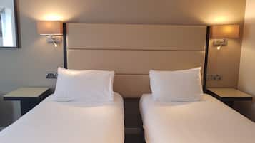 Quarto de hotel com duas camas de solteiro, cada uma com uma almofada e uma mesa de cabeceira. Há luminárias fixadas na parede, uma de cada lado das camas.