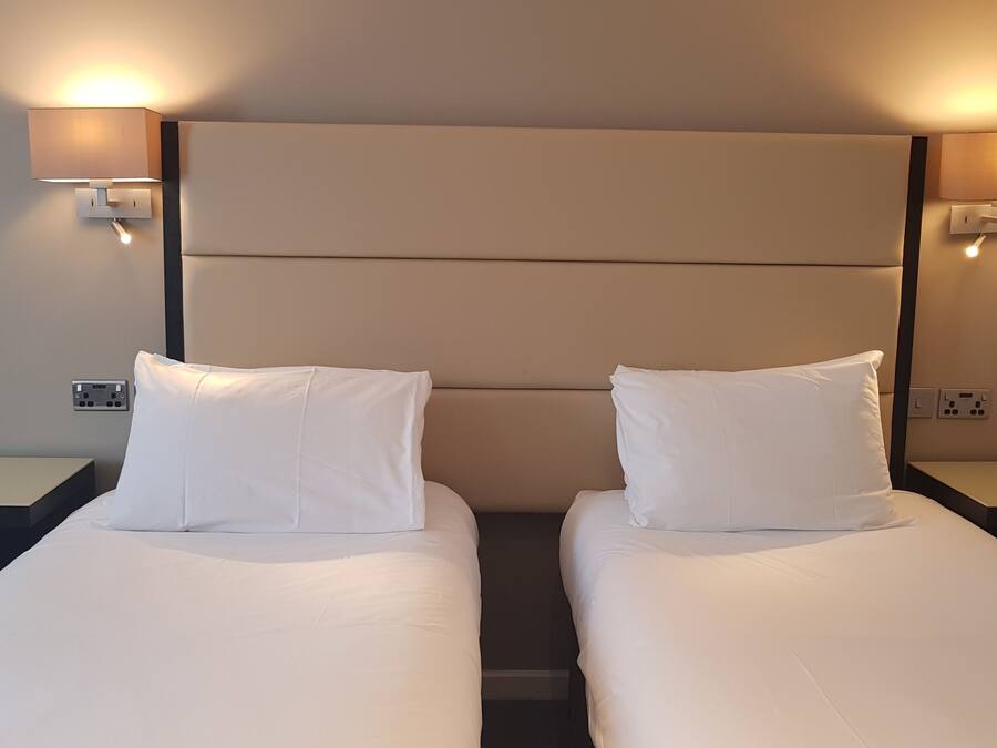 Quarto de hotel com duas camas de solteiro, cada uma com uma almofada e uma mesa de cabeceira. Há luminárias fixadas na parede, uma de cada lado das camas.