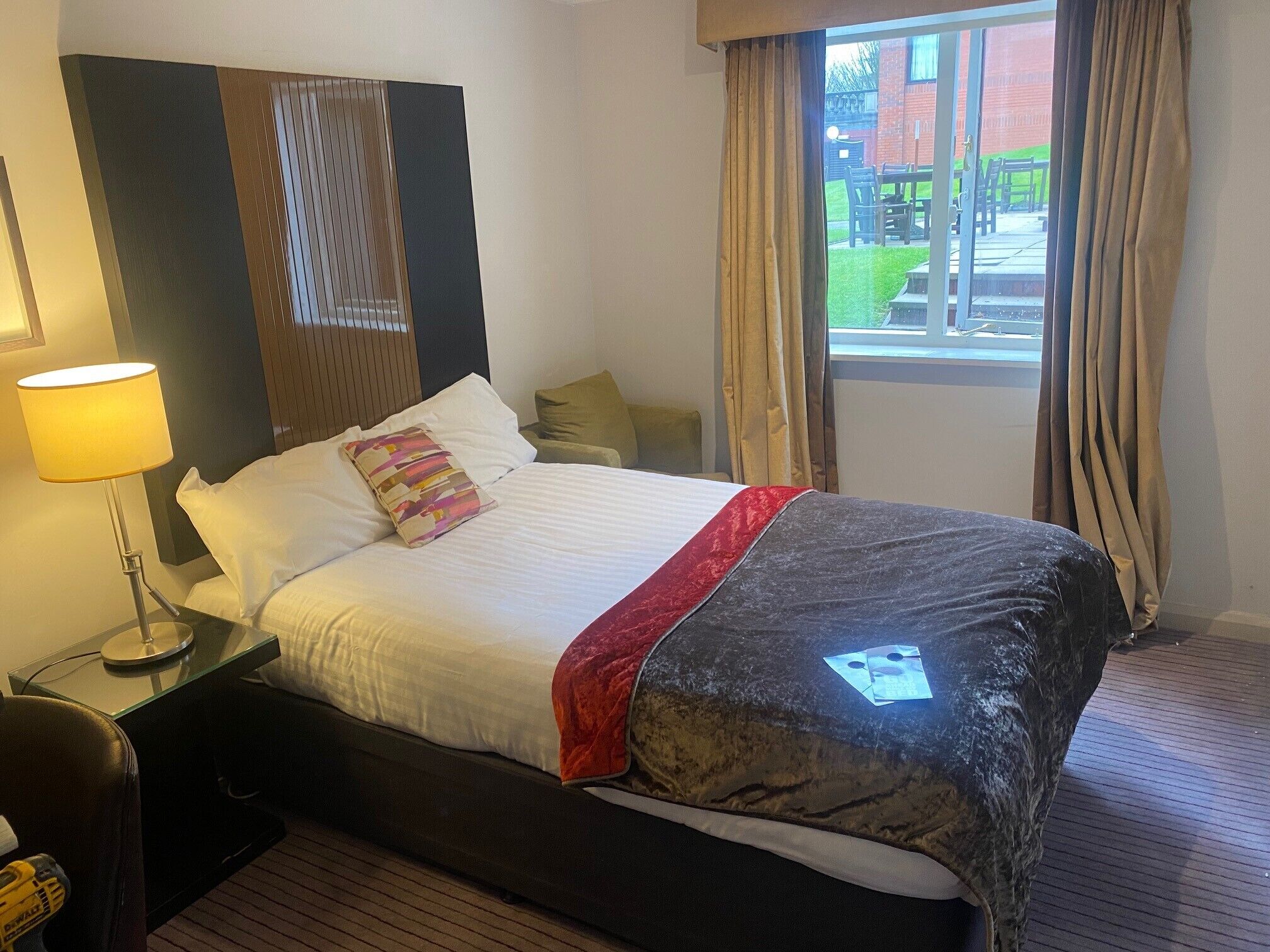 Quarto de hotel com cama, mesa de cabeceira, luminária e janela com cortinas.