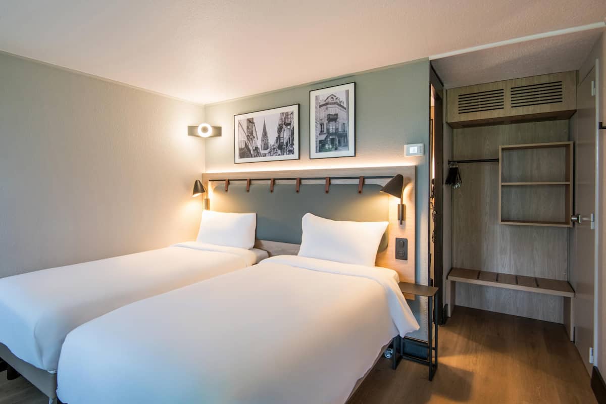 Quarto de hotel com duas camas, uma televisão fixada na parede, um cabideiro e um guarda-roupa de madeira.