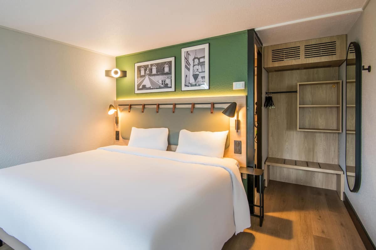 Quarto de hotel com uma cama, uma luminária fixada na parede, um cabideiro e um guarda-roupa de madeira.