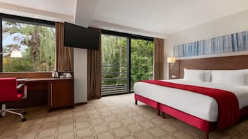 Quarto de hotel com uma cama grande, uma escrivaninha com cadeira, televisão e vista para árvores pela janela.