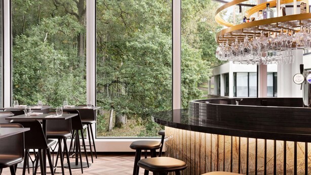 Uma área de bar moderna com balcão curvo, banquetas e vista para uma floresta através de grandes janelas.