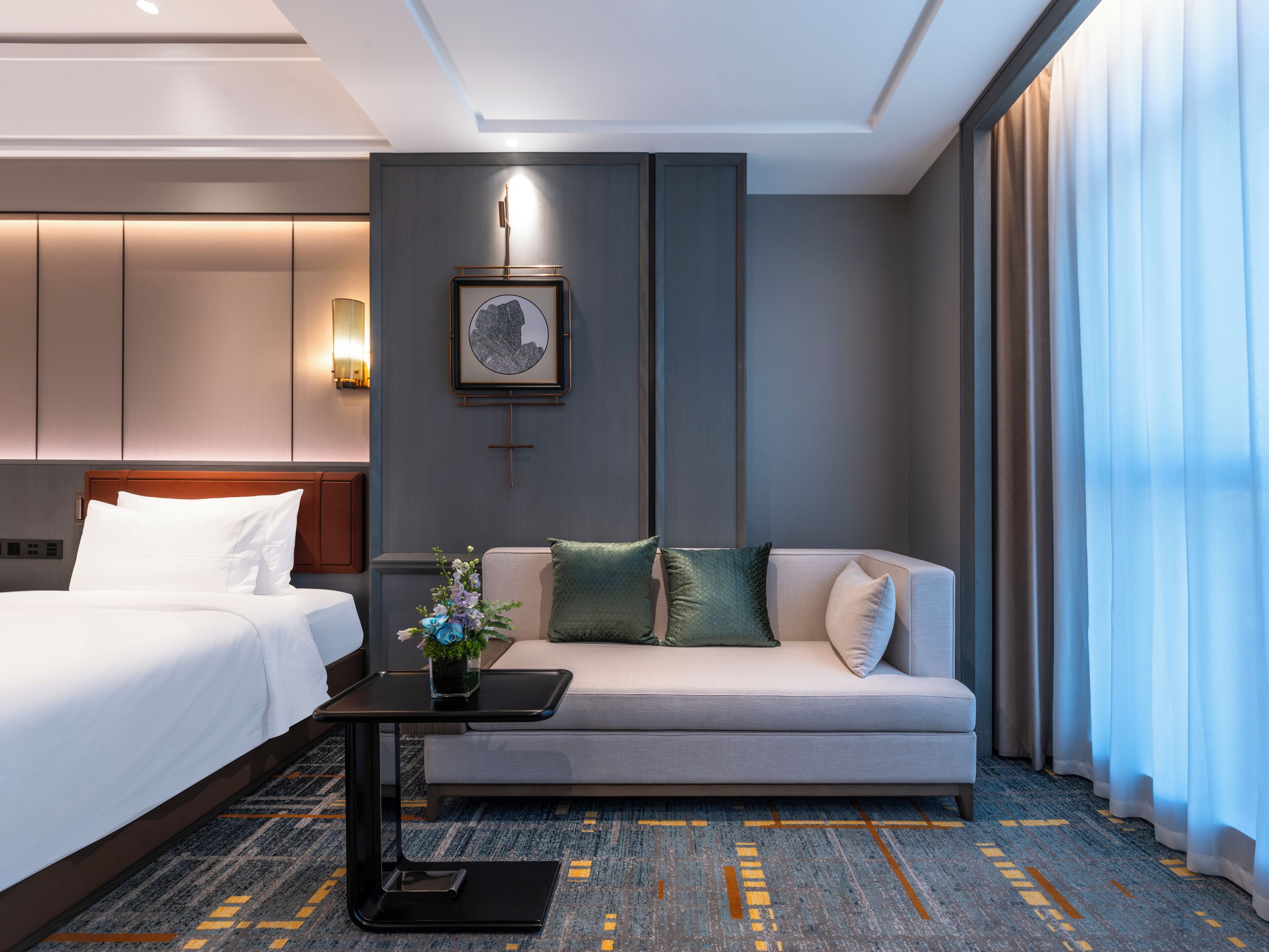 Quarto de hotel com cama, sofá, uma mesa pequena e uma obra de arte na parede.