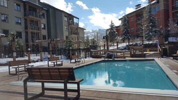 Piscina com um banco de madeira em primeiro plano, rodeada por edifícios e árvores cobertas de neve.