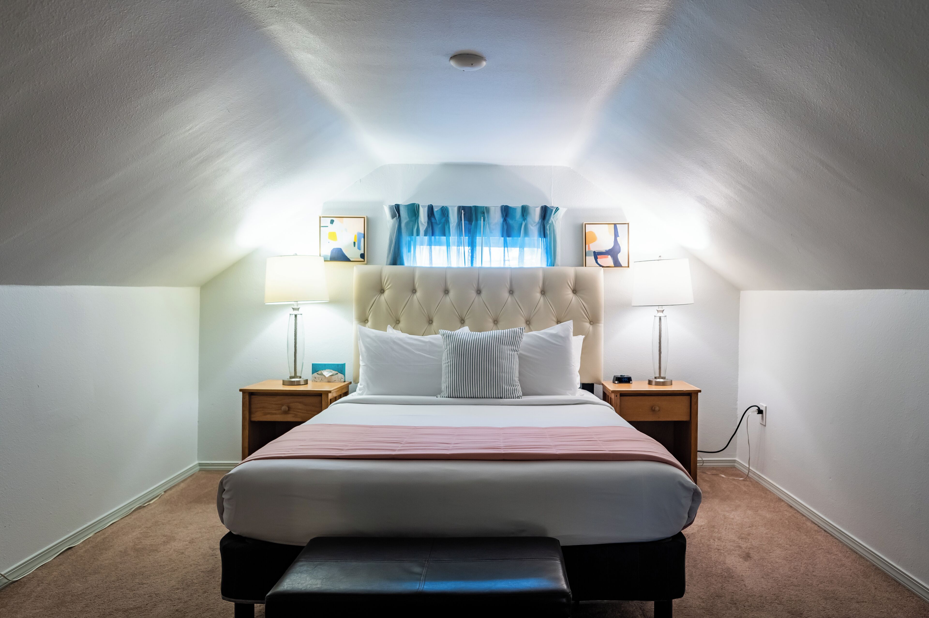 Quarto com cabeceira estofada, cama grande, luminárias de cabeceira e janela com cortinas azuis.