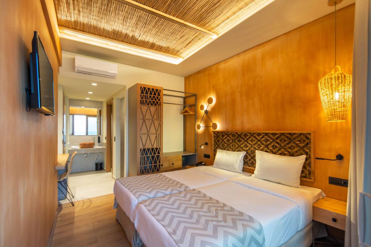 Quarto de hotel moderno com uma cama grande, cabeceira de madeira, TV de tela plana na parede e uma pequena cozinha no fundo.