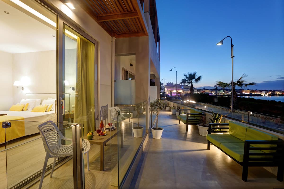Quarto de hotel moderno com varanda, área de jantar e vista para a paisagem urbana ao entardecer.
