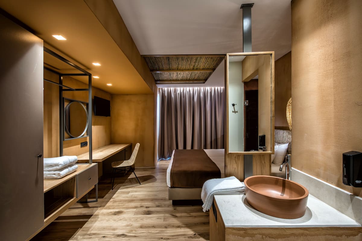 Quarto de hotel moderno com cama, escrivaninha de madeira, espelho grande, banheiro com pia e vaso sanitário, e área de chuveiro.