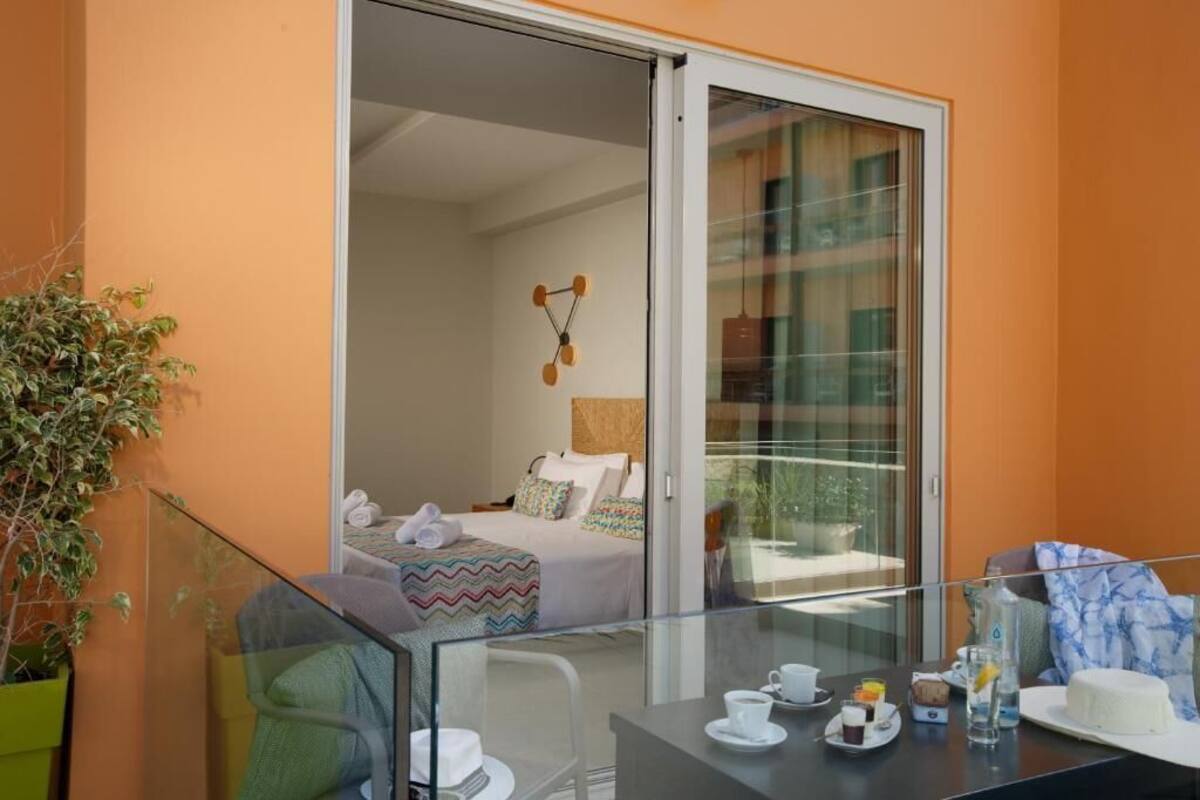 Varanda com mesa de vidro posta para o café da manhã, cama com colcha colorida e vista do interior do quarto.