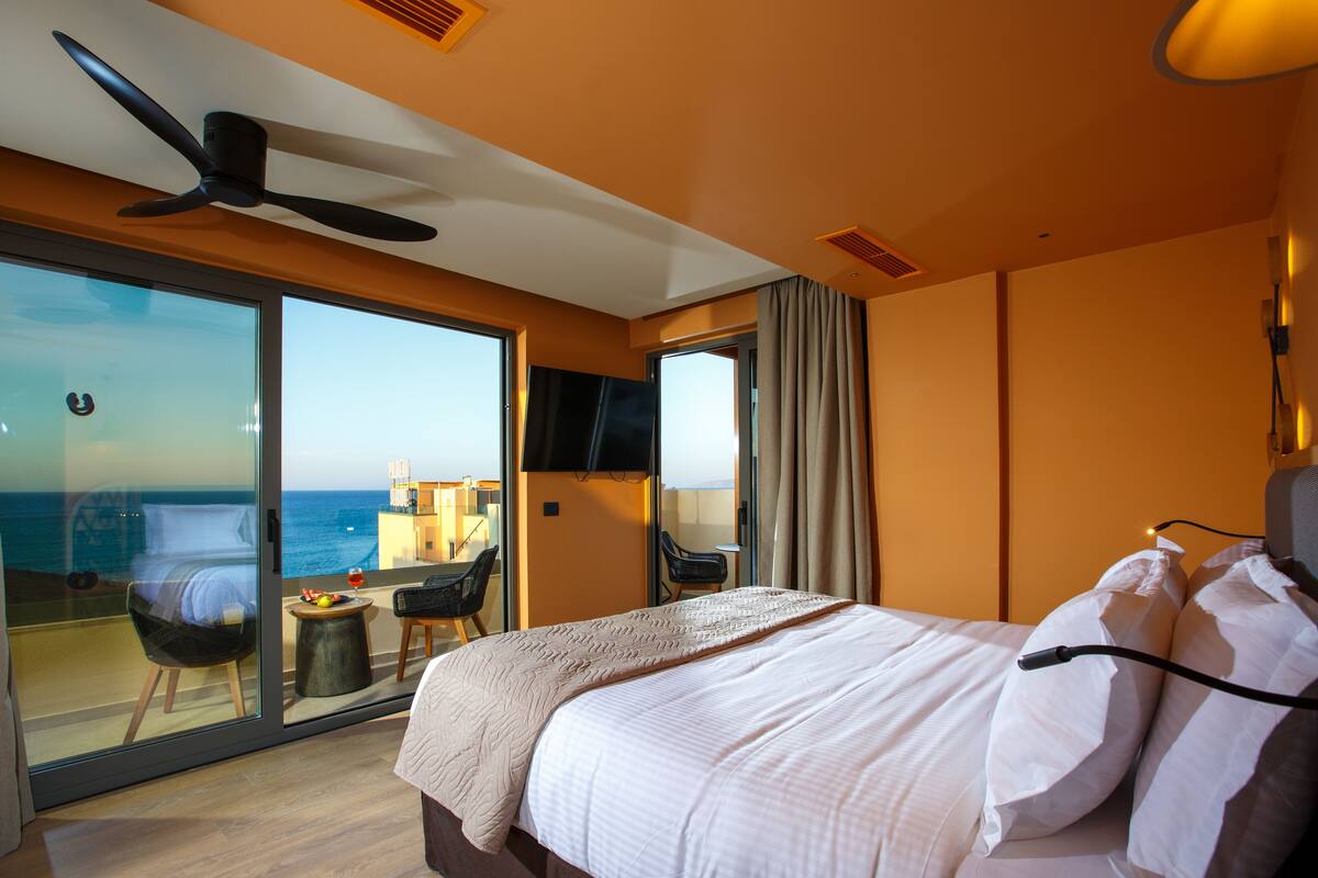 Quarto de hotel com cama, ventilador de teto, televisão, varanda com cadeira e vista para o mar.