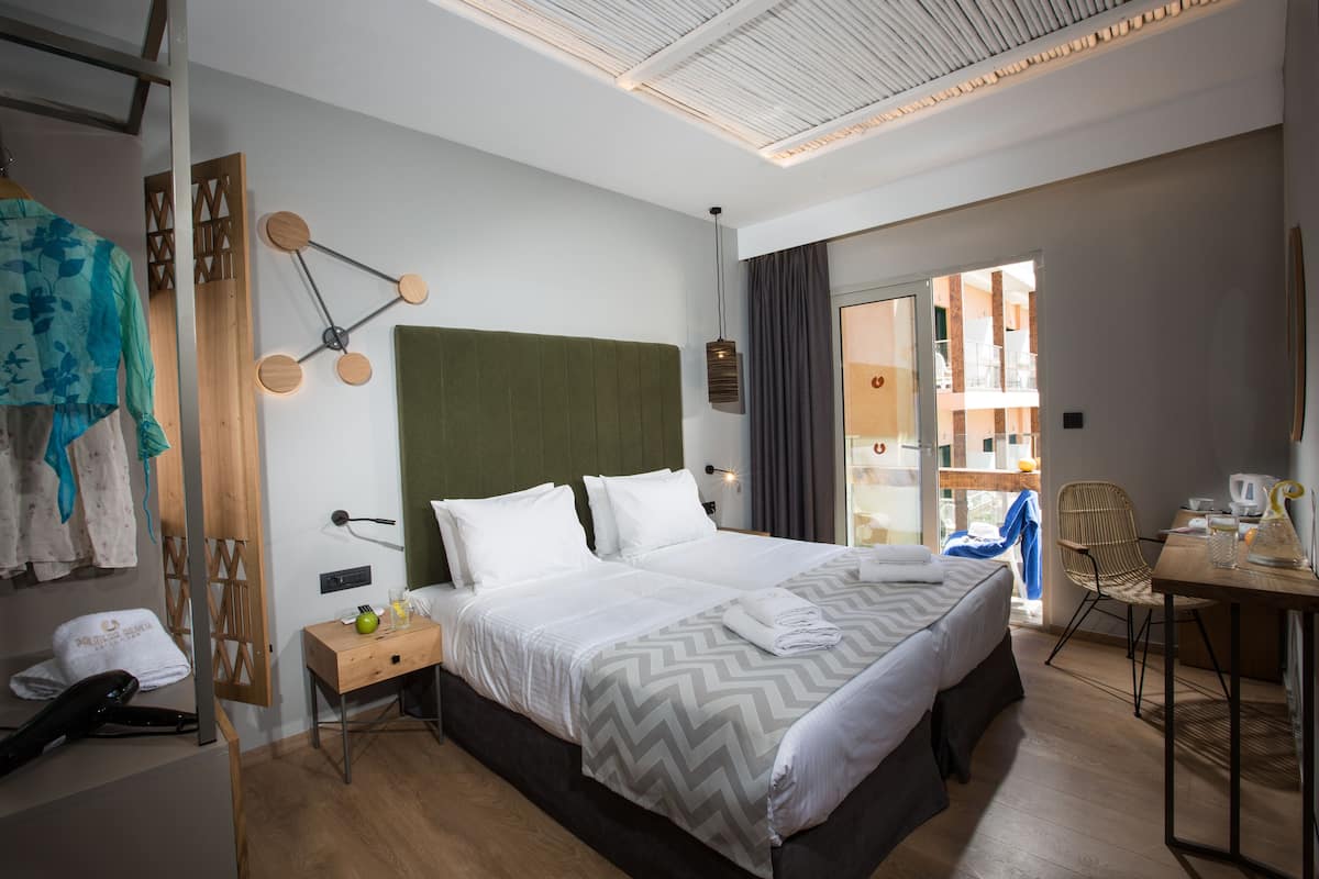 Quarto de hotel com uma cama grande, mesa de cabeceira de madeira, escrivaninha e uma cadeira. Há vista para o exterior através de uma porta de vidro.