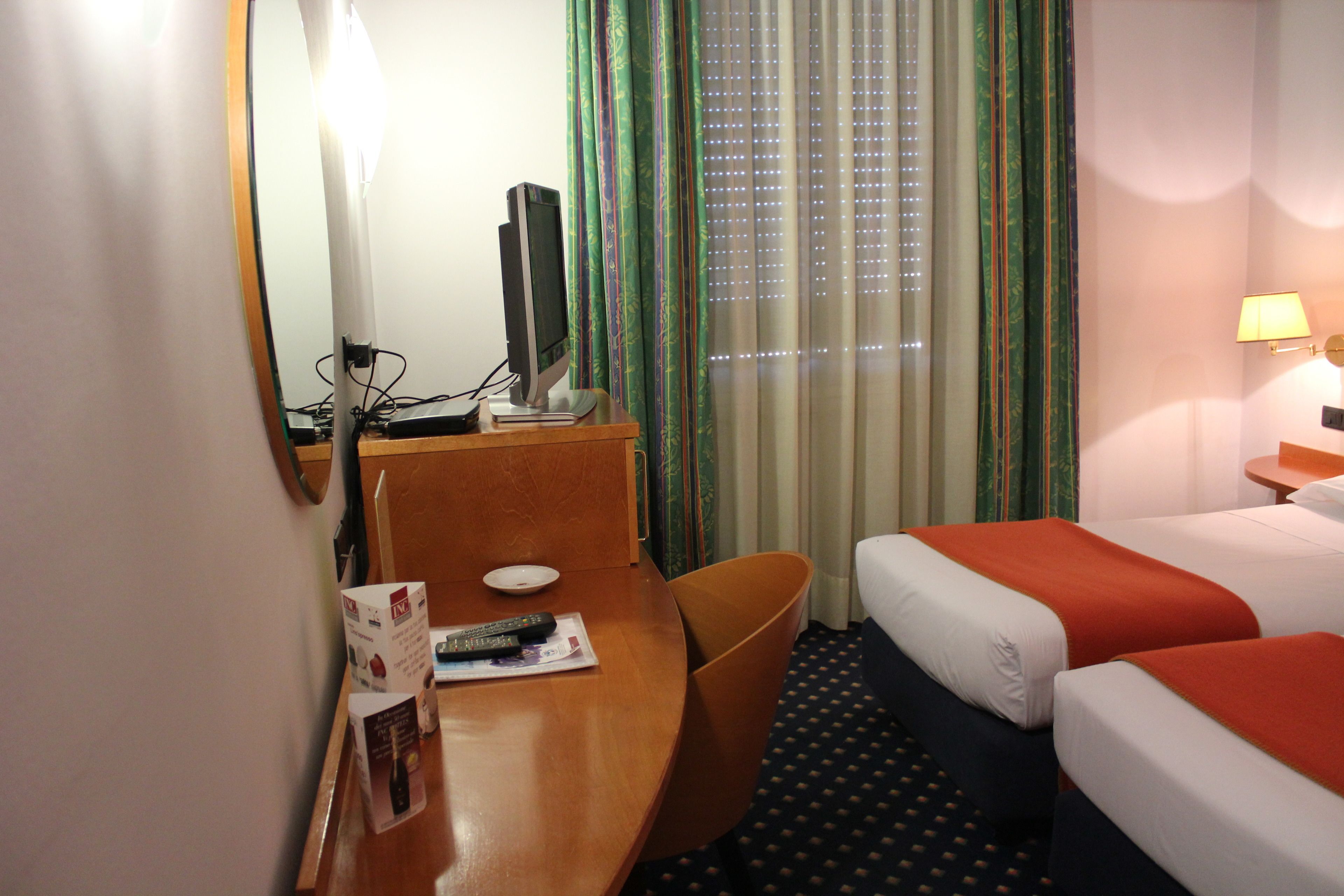 Quarto de hotel com uma escrivaninha, televisão, duas camas e uma janela com cortinas.