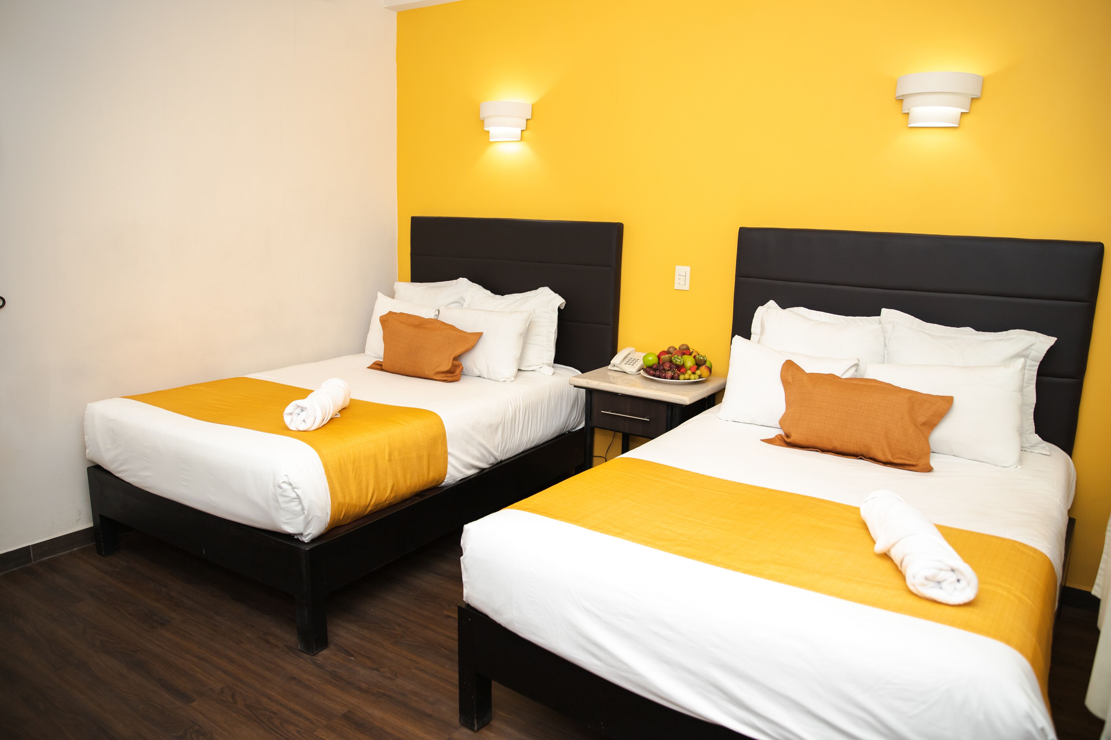 Duas camas de casal com roupa de cama branca e amarela, uma mesinha com fruta e luzes fixadas na parede.