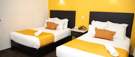 Duas camas de casal com roupa de cama branca e amarela, uma mesinha com fruta e luzes fixadas na parede.