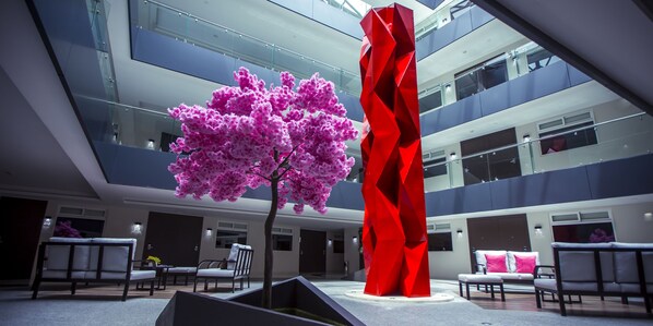 Espaço interior moderno com uma escultura vermelha central, uma árvore com flores cor-de-rosa e áreas de estar.