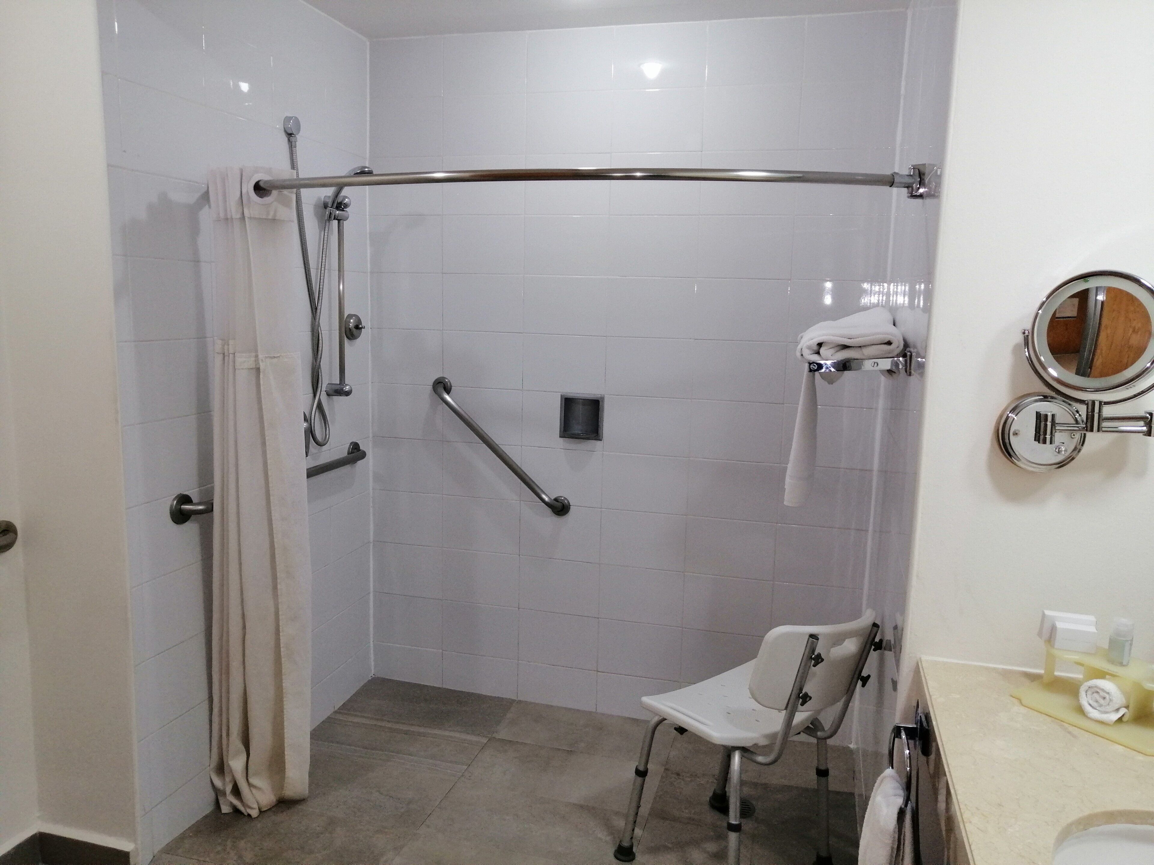 Casa de banho com chuveiro, barras de apoio, cadeira acessível para cadeira de rodas e espelho.