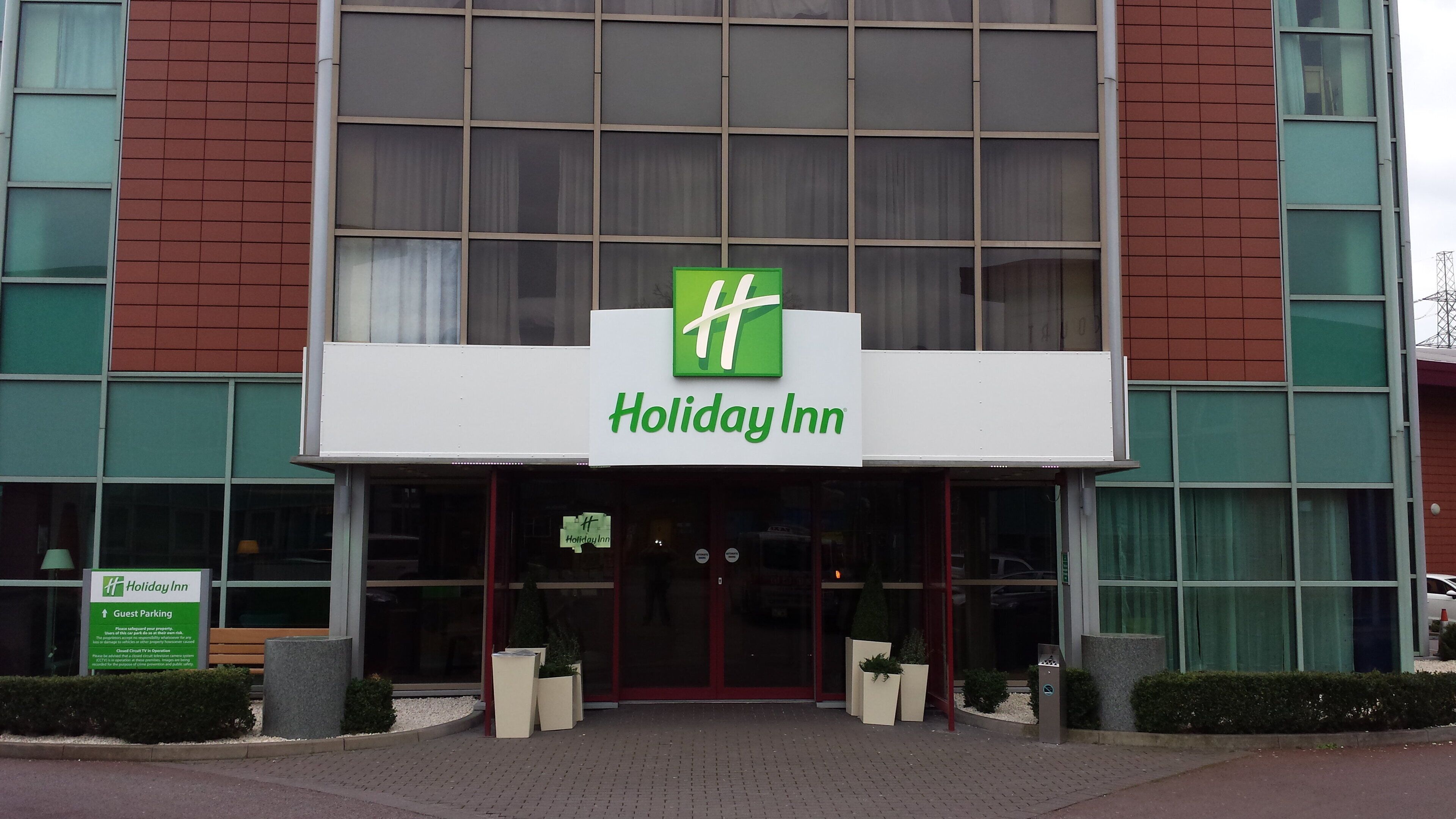 Entrada de um hotel Holiday Inn com um logotipo verde bem visível, placa indicando estacionamento para hóspedes e fachada moderna com grandes janelas.