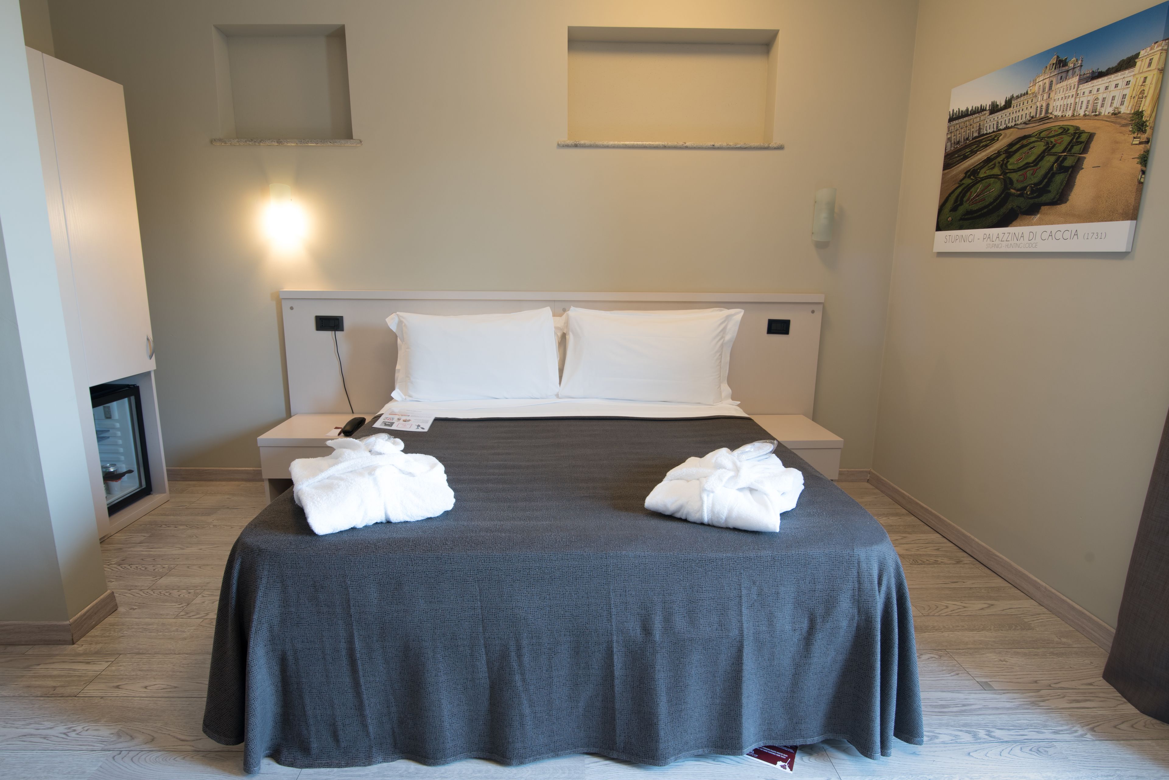 Quarto de hotel com uma cama grande, duas almofadas brancas, um edredom cinzento e duas toalhas sobre a cama. Há uma pequena mesa de cabeceira com um telefone e uma luz na parede.