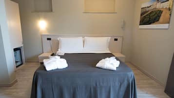 Quarto de hotel com uma cama grande, duas almofadas brancas, um edredom cinzento e duas toalhas sobre a cama. Há uma pequena mesa de cabeceira com um telefone e uma luz na parede.