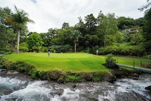 Campo de golfe com um rio em primeiro plano, cercado por vegetação exuberante e árvores.