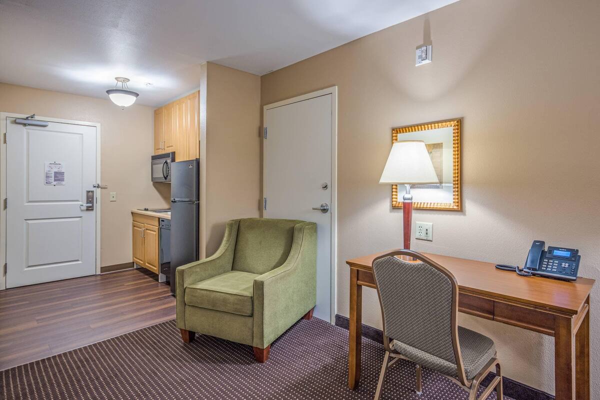 Quarto de hotel com cozinha compacta, uma poltrona verde, uma escrivaninha com cadeira, luminária e telefone.