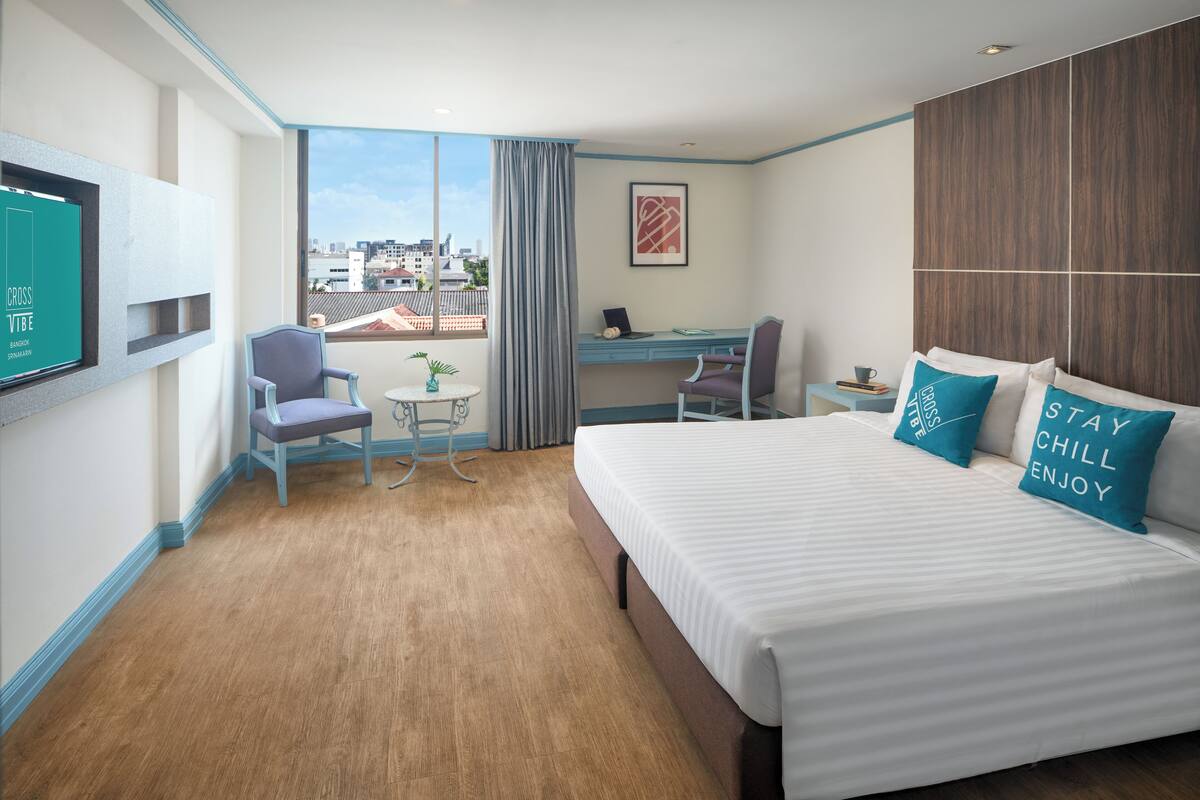 Quarto de hotel com uma cama grande, uma escrivaninha, uma cadeira, uma mesa pequena e vista da cidade através de janelas amplas.