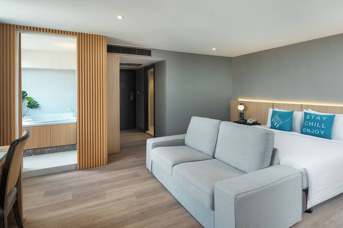 Quarto de hotel moderno com sofá cinza, cama com almofadas verde-água, divisória de madeira e vista para uma área de spa.