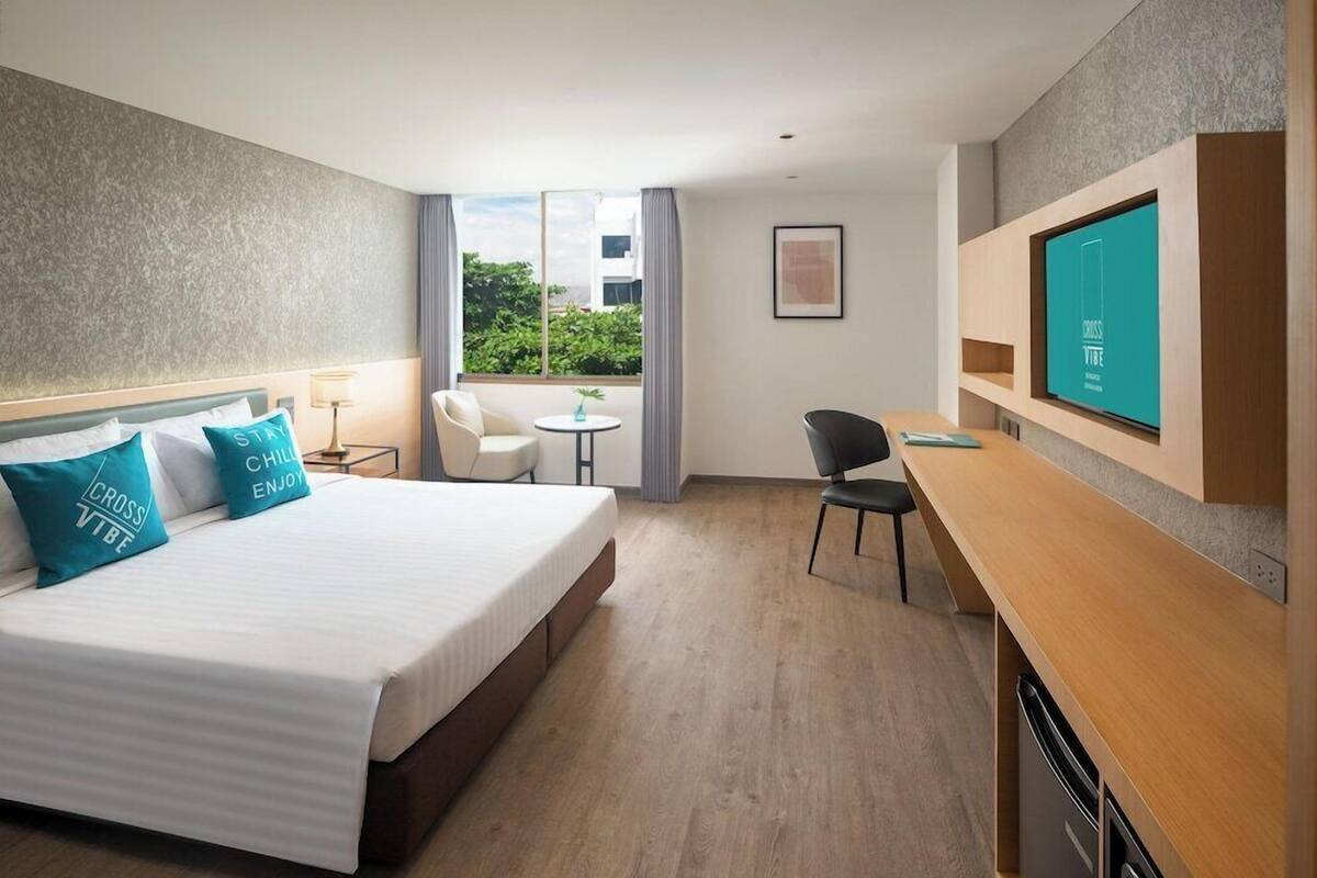 Quarto de hotel com cama, escrivaninha, cadeira, mesa pequena, televisão e janela com vista para edifícios e vegetação.