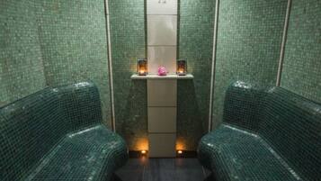 Casa de banho com azulejos de mosaico verdes, duas pias e um armário central com velas e uma flor rosa.