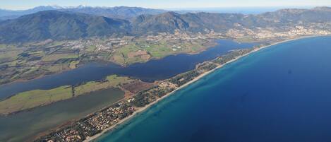 Vista aérea de uma cidade costeira com uma praia, montanhas ao fundo e uma grande extensão de água.