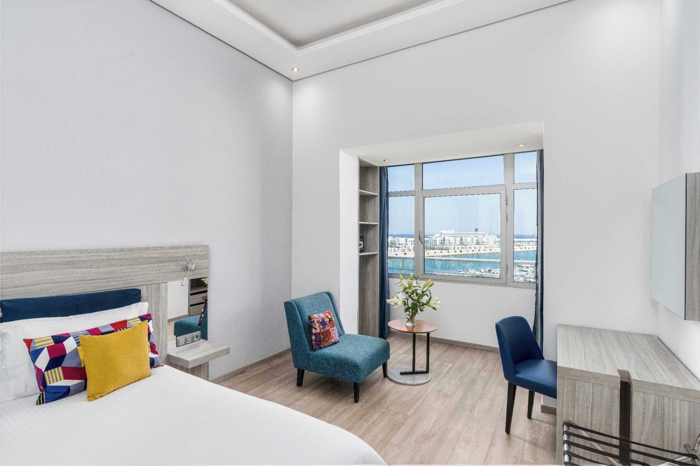 Quarto de hotel moderno com uma cama grande, escrivaninha, cadeira e janela com vista para a cidade.