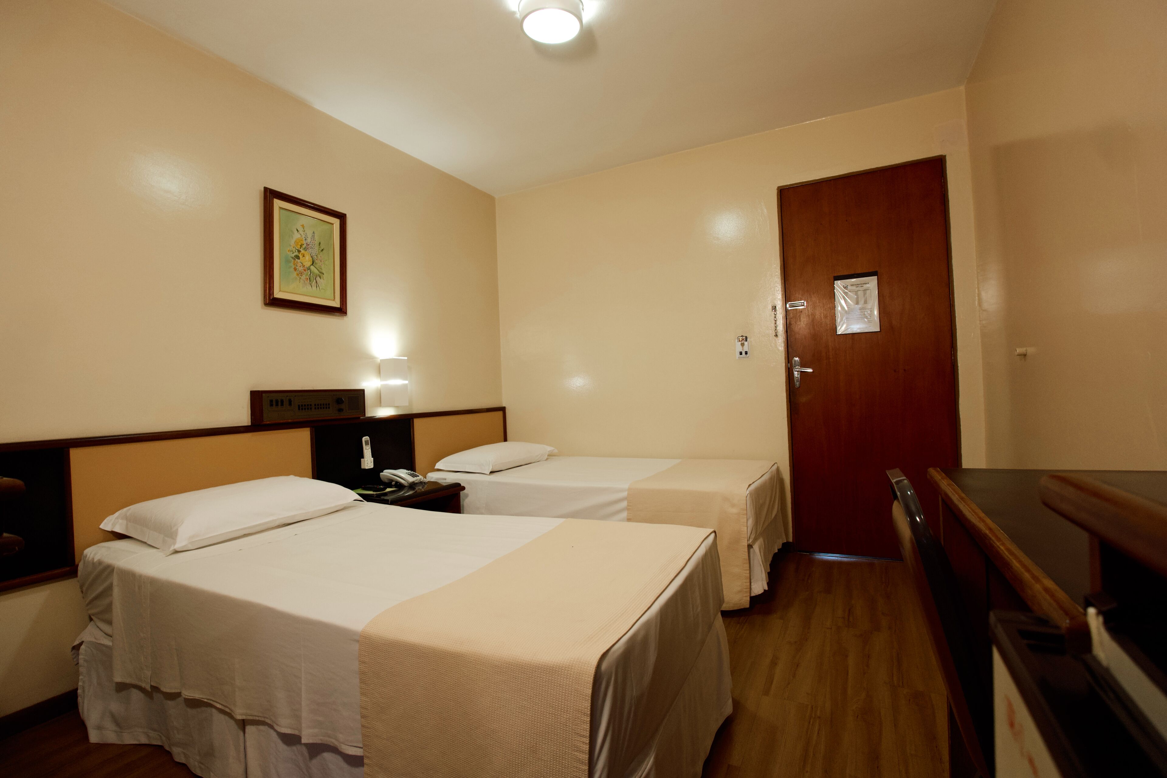 Quarto de hotel com duas camas, penteadeira de madeira, mesa de cabeceira, quadro na parede e porta com uma placa.