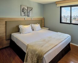 Quarto de hotel com cama grande, cabeceira, janela com vista para a cidade e duas obras de arte penduradas na parede.
