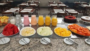 Buffet com diversas frutas, sumos e um bolo em área de refeições com mesas e cadeiras.