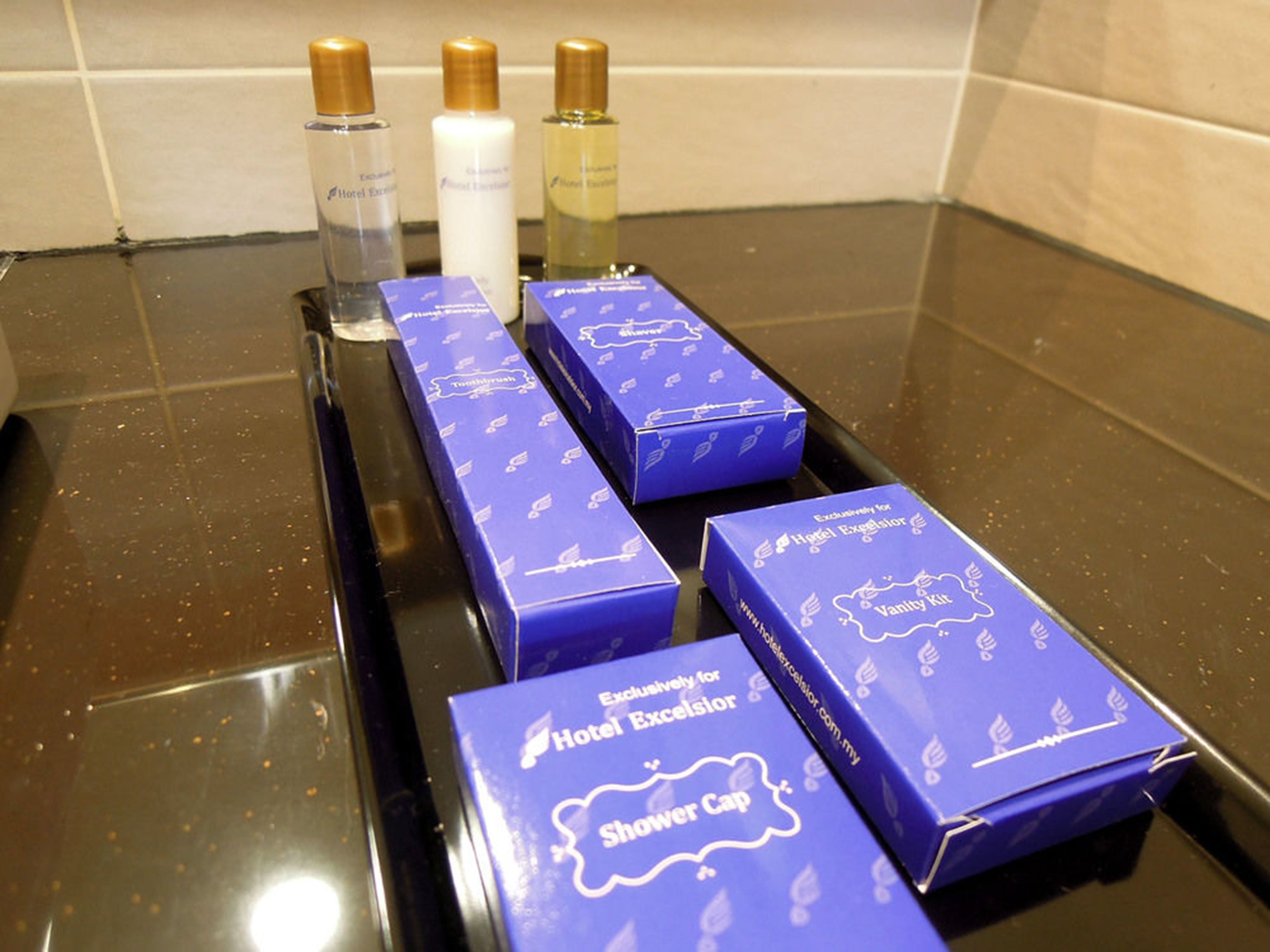 Uma bancada de banheiro com quatro caixas azuis identificadas como “Hotel Excelsior Shower Cap” e três frascos de artigos de higiene pessoal.