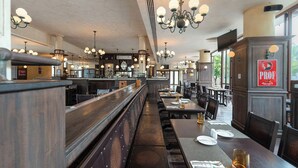 Interior de restaurante com mesas comunitárias alongadas, área de bar e lustres pendurados.