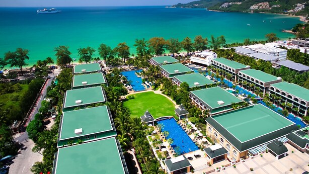 Um resort com vários edifícios com telhados verdes, uma piscina central e vista para o oceano e colinas.