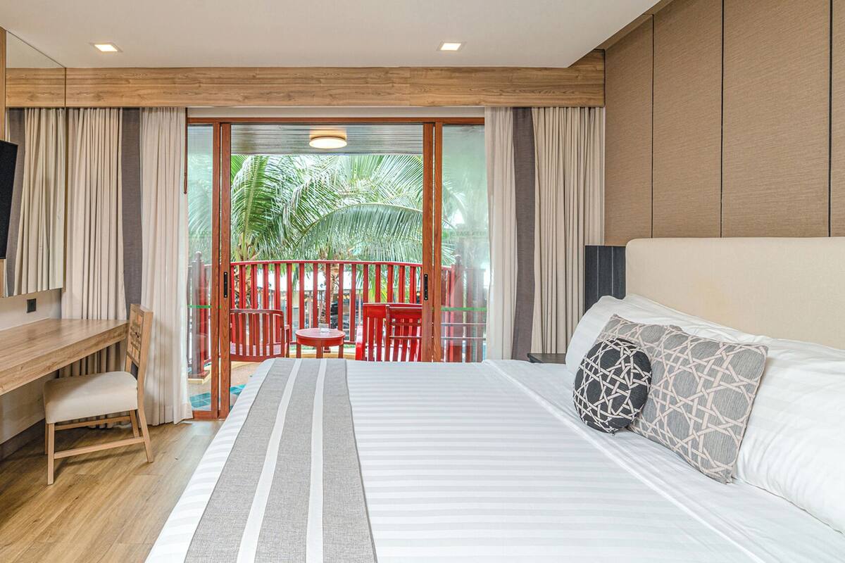 Quarto de hotel com cama, escrivaninha, televisão e varanda com cadeiras vermelhas.