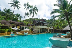 Área de piscina de resort com água azul cristalina, cadeiras de lounge, guarda-sóis e palmeiras.