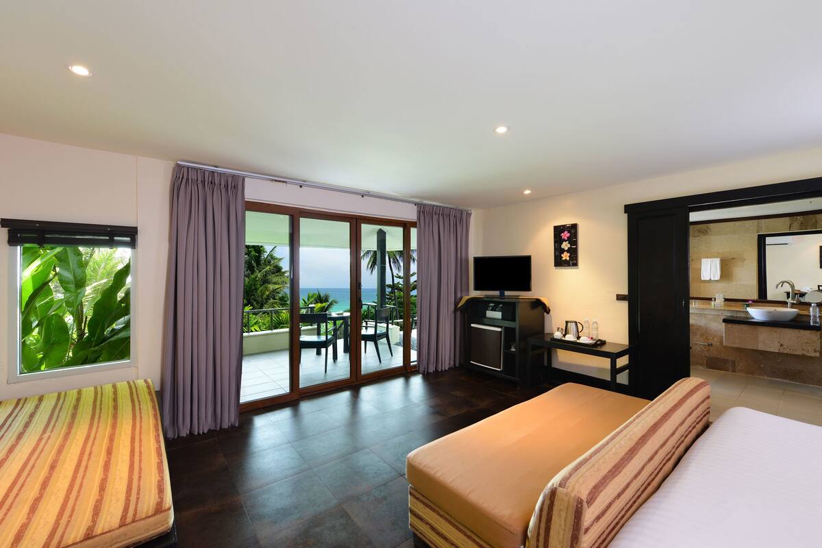 Quarto de hotel com cama, escrivaninha com televisão, banheiro com pia e vista para o oceano através de grandes janelas.