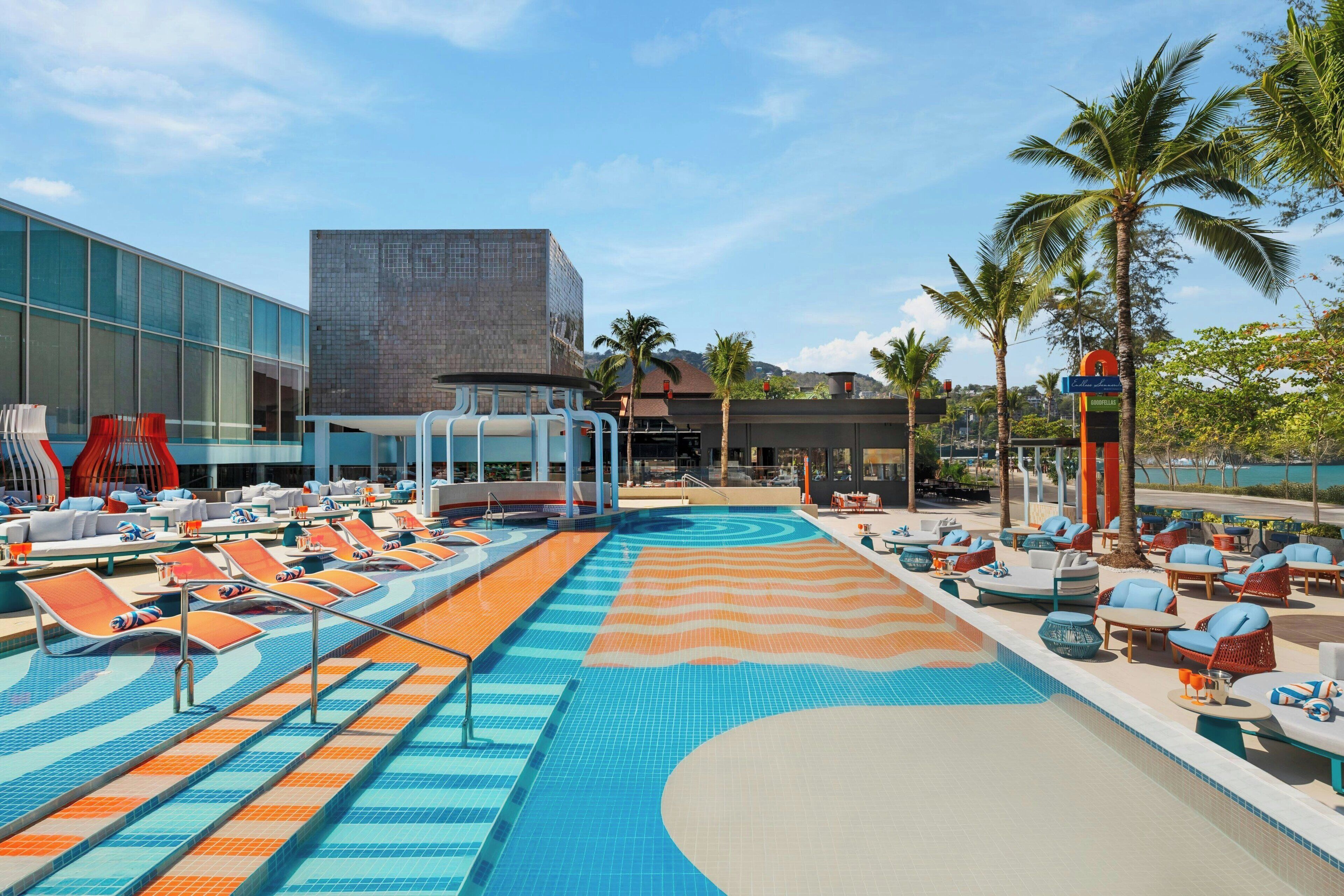 Área de piscina com espreguiçadeiras e mesas, um edifício moderno com amplas janelas e palmeiras ao fundo.