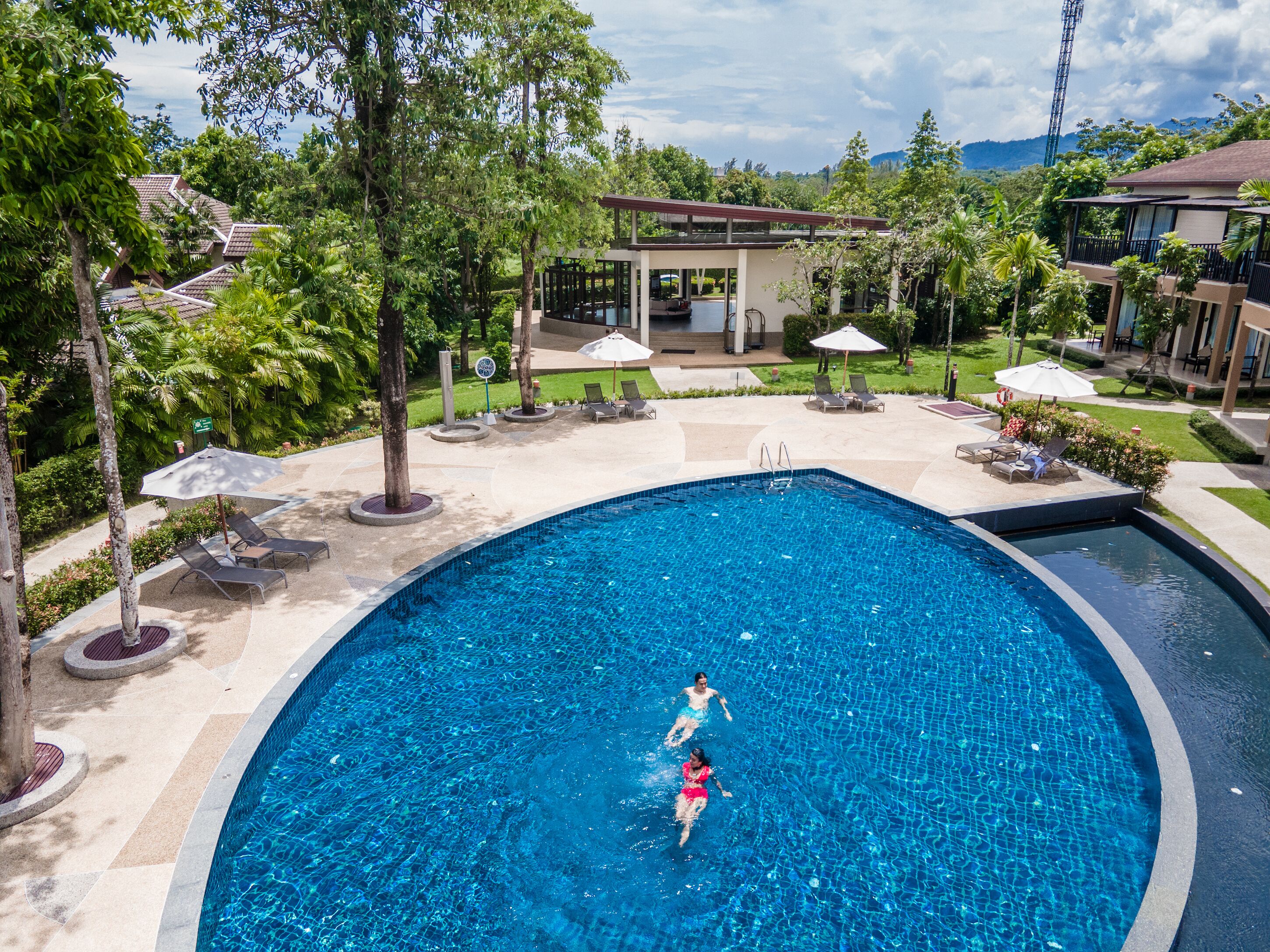 Uma grande piscina ao ar livre rodeada por espreguiçadeiras e guarda-sóis, com vista para um edifício e árvores ao fundo.