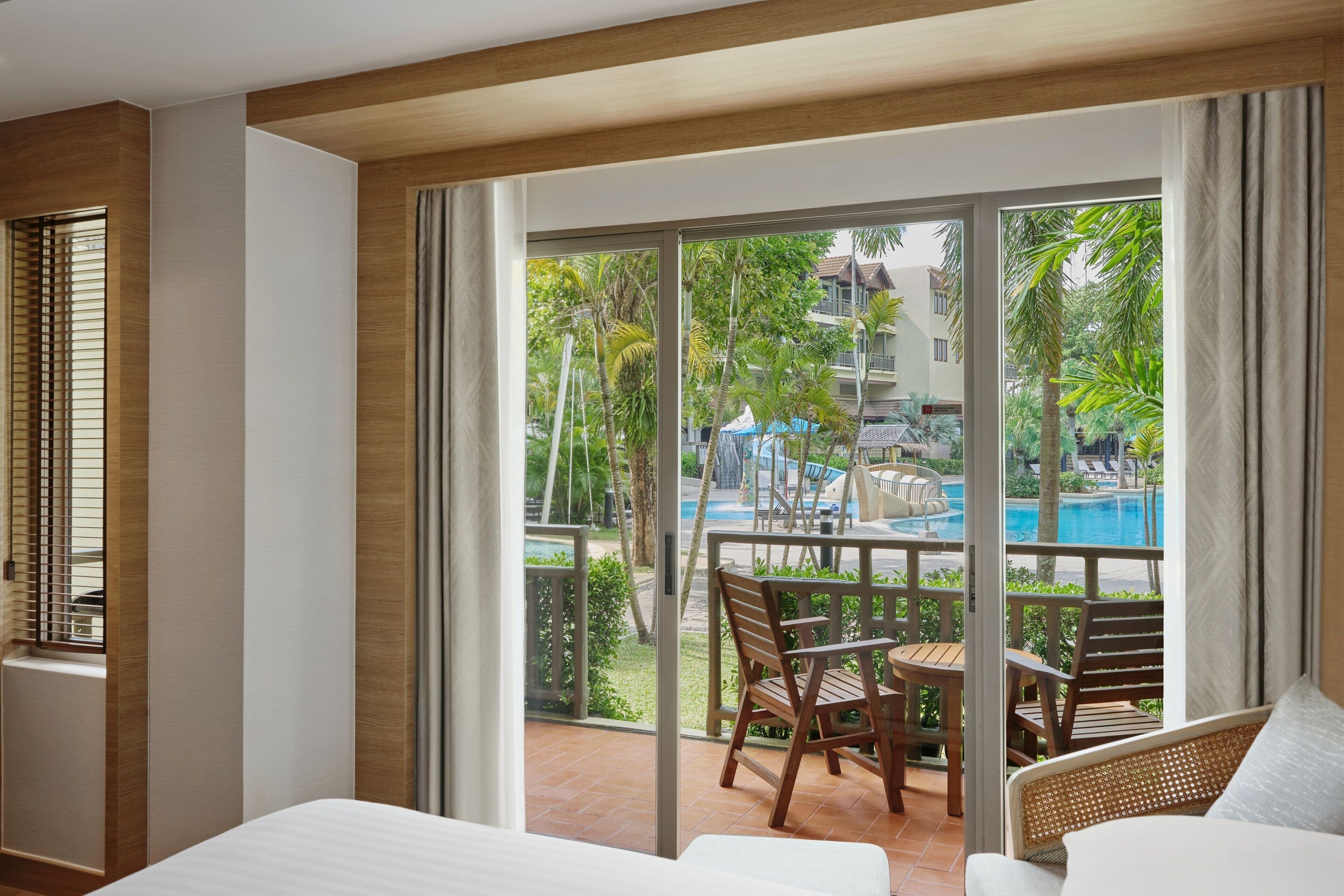 Quarto de hotel com uma grande porta de vidro deslizante que dá para uma área de piscina, mobiliário de madeira e vista para a piscina e vegetação ao redor.