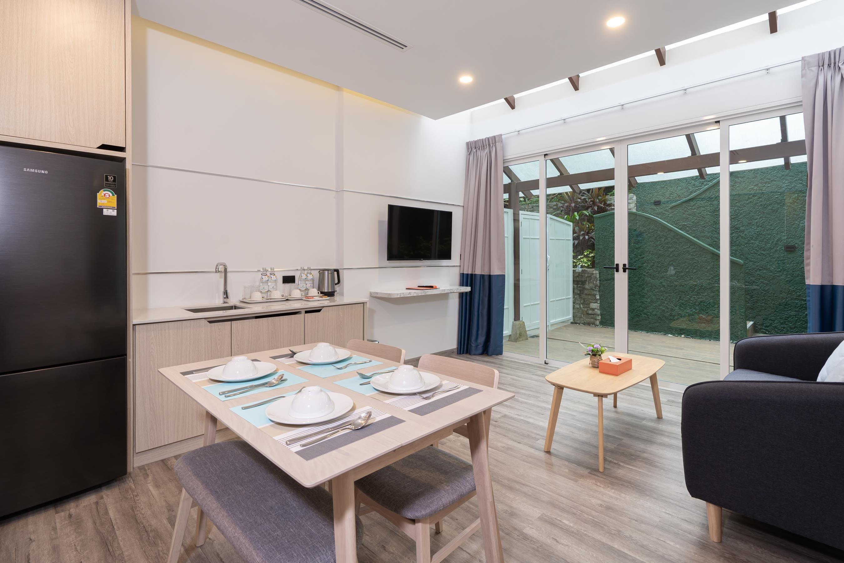 Uma cozinha moderna com geladeira Samsung embutida, sala de jantar com mesa posta para quatro pessoas e sala de estar com sofá e TV.