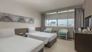 Quarto de hotel com duas camas, uma escrivaninha, uma cadeira e uma janela grande com vista para a cidade.